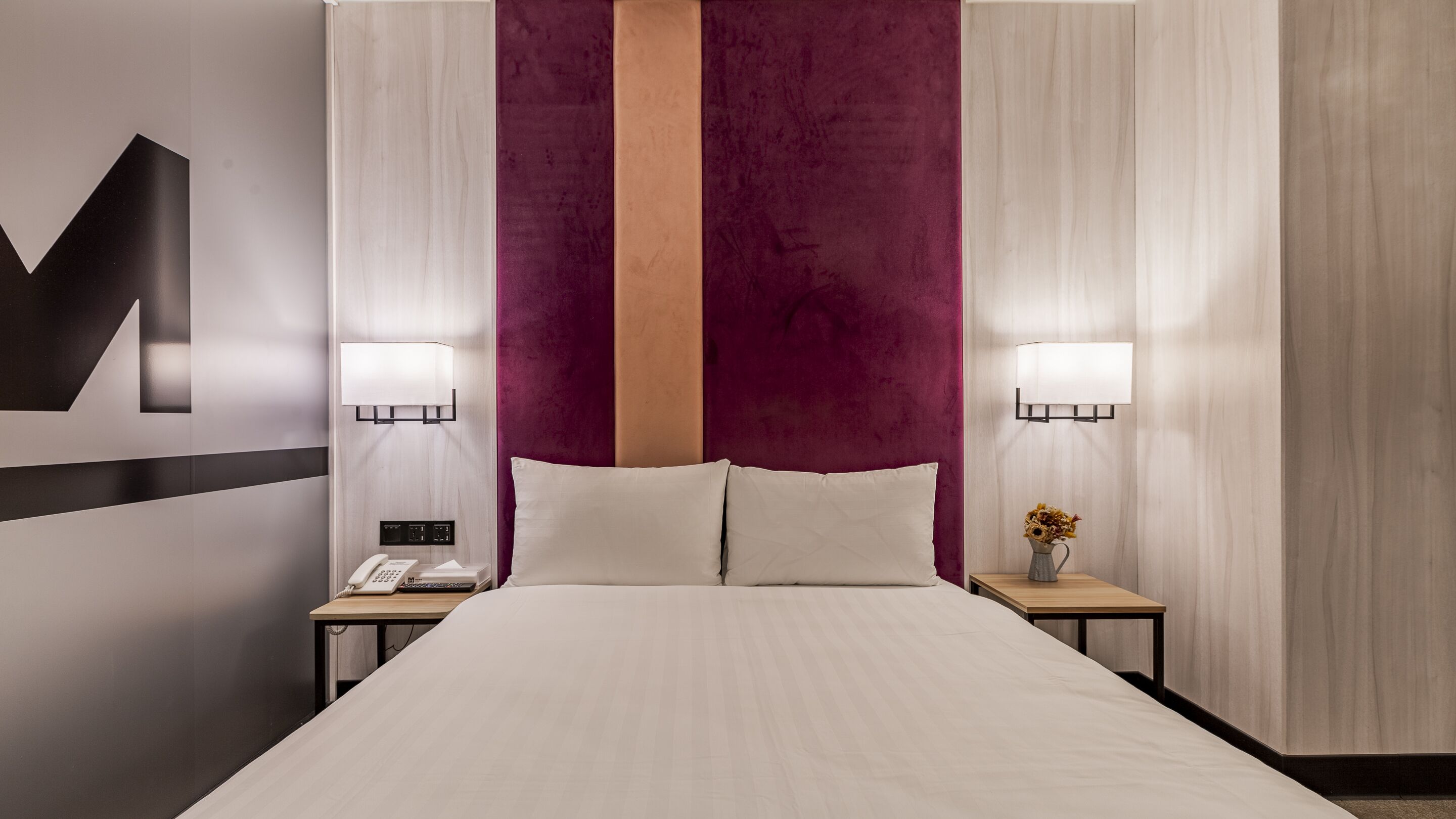 Quarto de hotel com uma cama grande, duas mesas de cabeceira, um telefone e duas luminárias fixadas na parede.