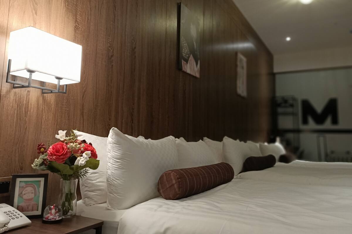 Quarto de hotel com parede de madeira, cama com almofadas brancas e castanhas, um vaso com flores, um quadro emoldurado e uma mesa de cabeceira com um telefone e um objeto decorativo.