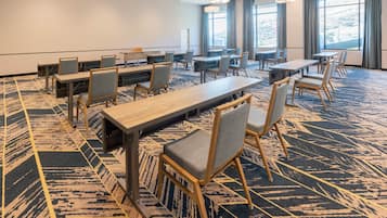 Uma sala de conferências com várias mesas e cadeiras, piso coberto de carpete e grandes janelas com cortinas.