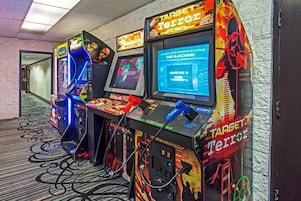 Fileira de máquinas de jogos de fliperama com a marca 'Target Terror', ligadas a uma fonte de energia, em um corredor com carpete estampado.