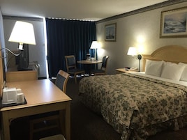 Quarto de hotel com uma cama, duas luminárias de cabeceira, uma escrivaninha com luminária, uma cadeira, uma mesinha com cadeira e um quadro na parede.