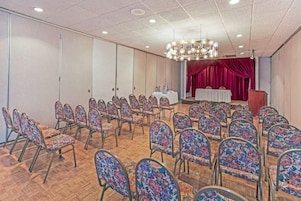 Uma sala de conferências com fileiras de cadeiras florais, um palco com cortina vermelha e um lustre.
