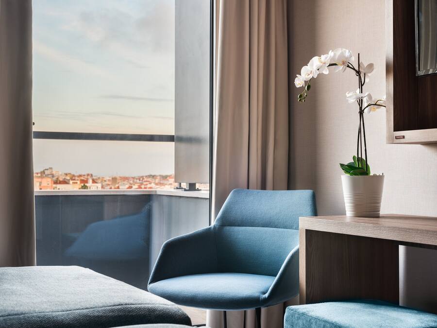 Quarto de hotel moderno com uma grande janela que oferece vista da cidade, uma poltrona azul, uma pequena mesa com uma planta em vaso e uma televisão de tela plana fixada na parede.