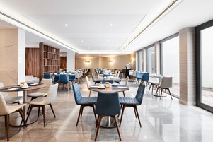 Ampla sala de jantar de hotel, com várias mesas e cadeiras, janelas grandes e design de interiores moderno.