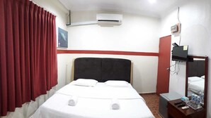 Quarto de hotel com cama, escrivaninha, ar condicionado, espelho e cortina vermelha.