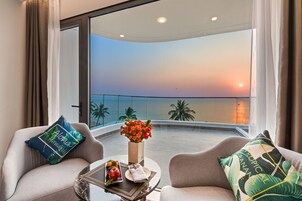 Uma varanda com vista para o pôr do sol, uma mesa com frutas e flores, e dois sofás com almofadas tropicais.