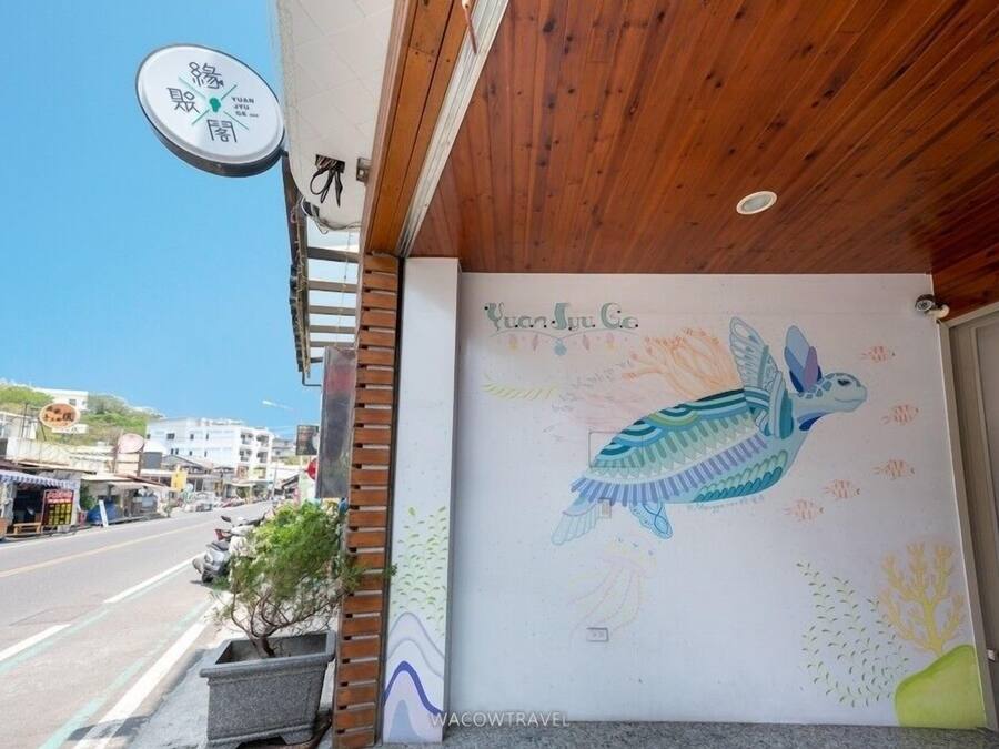 Um edifício com um mural de tartaruga marinha, uma placa com caracteres chineses e uma vista da rua com lojas e céu claro.