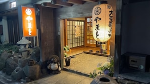 Uma entrada tradicional japonesa com um candeeiro de pedra, uma porta de madeira e um lança-luz com caracteres japoneses.