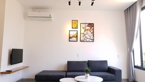 Uma sala de estar moderna com um sofá cinza escuro, duas mesinhas de apoio brancas e uma TV de tela plana em uma estante de madeira. Há três quadros na parede e um ar condicionado acima do sofá.