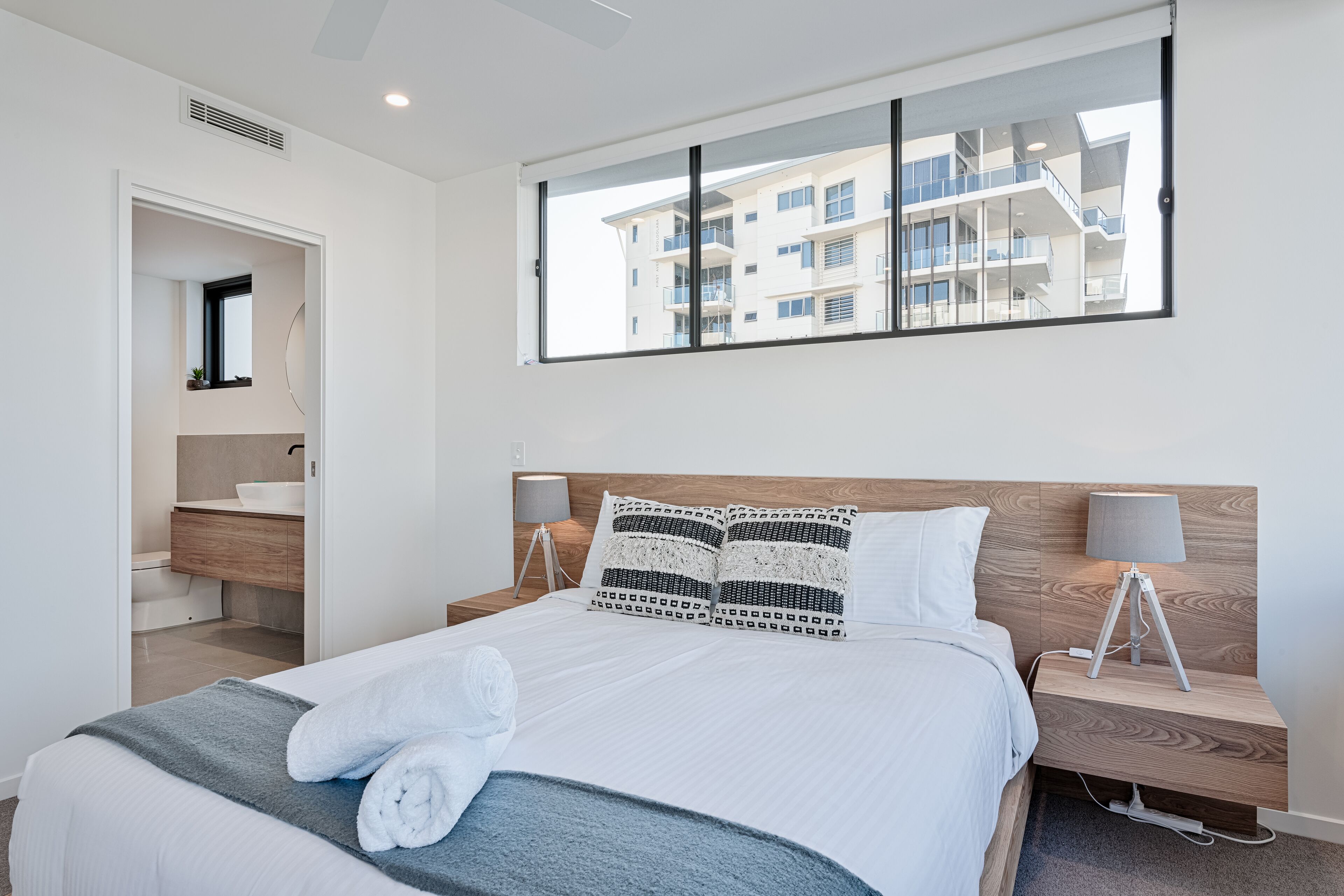 Quarto de hotel moderno com uma cama grande, cabeceira de madeira, duas luminárias de apoio e vista para um edifício alto pela janela.