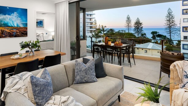 Uma sala de estar moderna com sofá, mesa de jantar e cadeiras. Há uma varanda com vista para o oceano e edifícios.