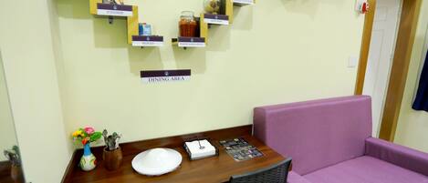 Uma área de refeições com uma mesa de madeira, cadeiras e um sofá roxo. Há prateleiras acima da mesa com diversos objetos e uma placa indicando a área de jantar.