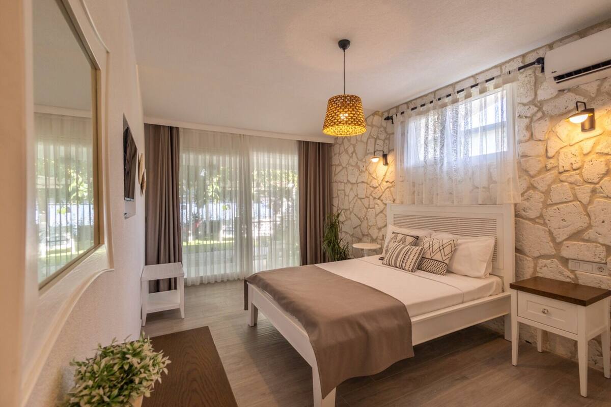 Quarto com parede de pedra, cama com almofadas estampadas, mesa de cabeceira e luminária suspensa.