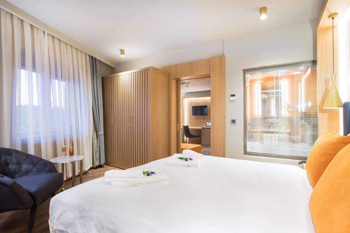 Quarto de hotel com uma cama grande, um espaço de estar com uma cadeira, uma mesinha com um copo e vista pela janela.