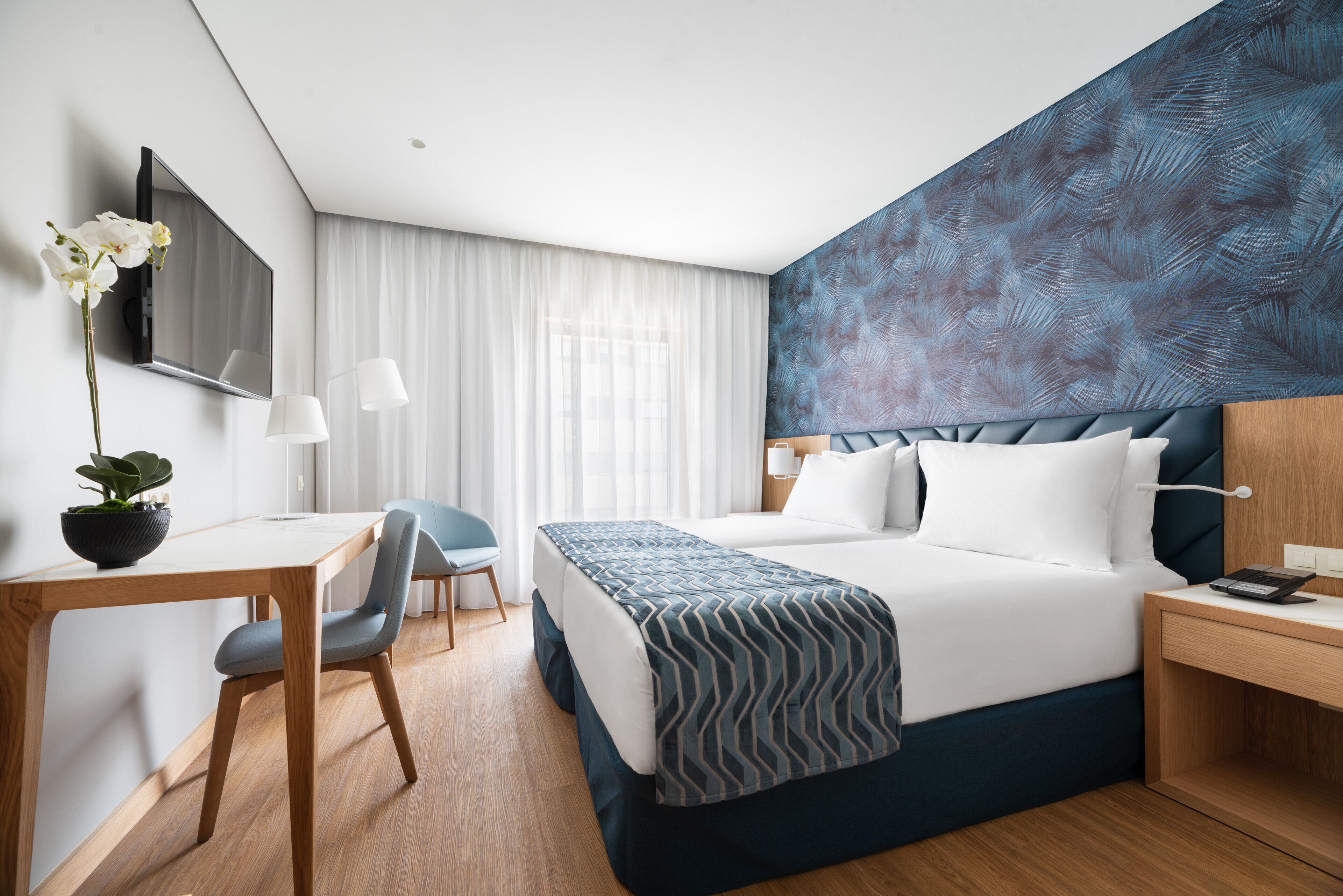 Quarto de hotel moderno com cama, mesa de cabeceira, luminária, televisão e escrivaninha com cadeira.