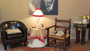 Um quarto com uma poltrona de couro preto, uma cadeira de madeira com almofada, uma mesa pequena com abajur, um quadro de um cavalo e uma mala florida.