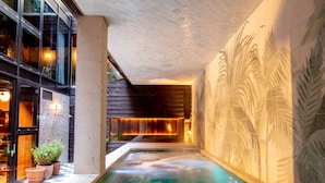 Uma área moderna de piscina interna, com um recurso aquático transparente, uma parede de vidro de um lado e outra parede com desenhos de palmeiras.