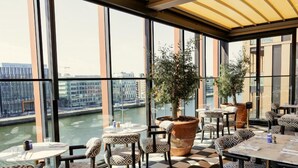 Um terraço no terraço com cadeiras e mesas estampadas, grandes janelas com vista para um rio e edifícios, e plantas em vasos.