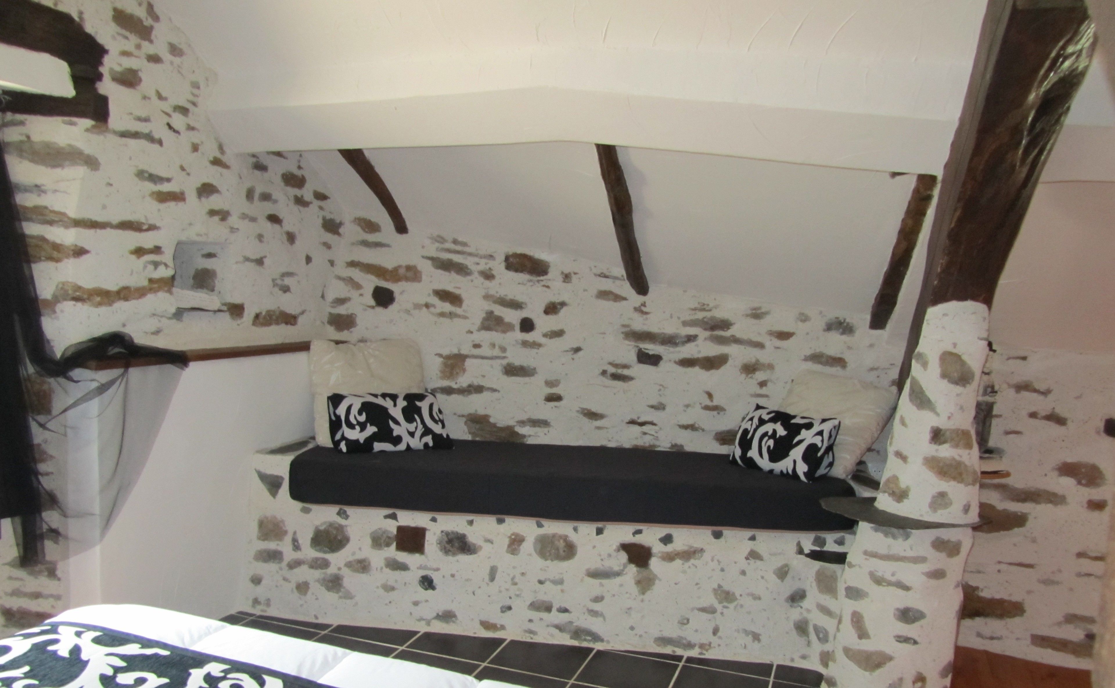 Quarto com parede de pedra, cama com cabeceira preta e duas almofadas estampadas.