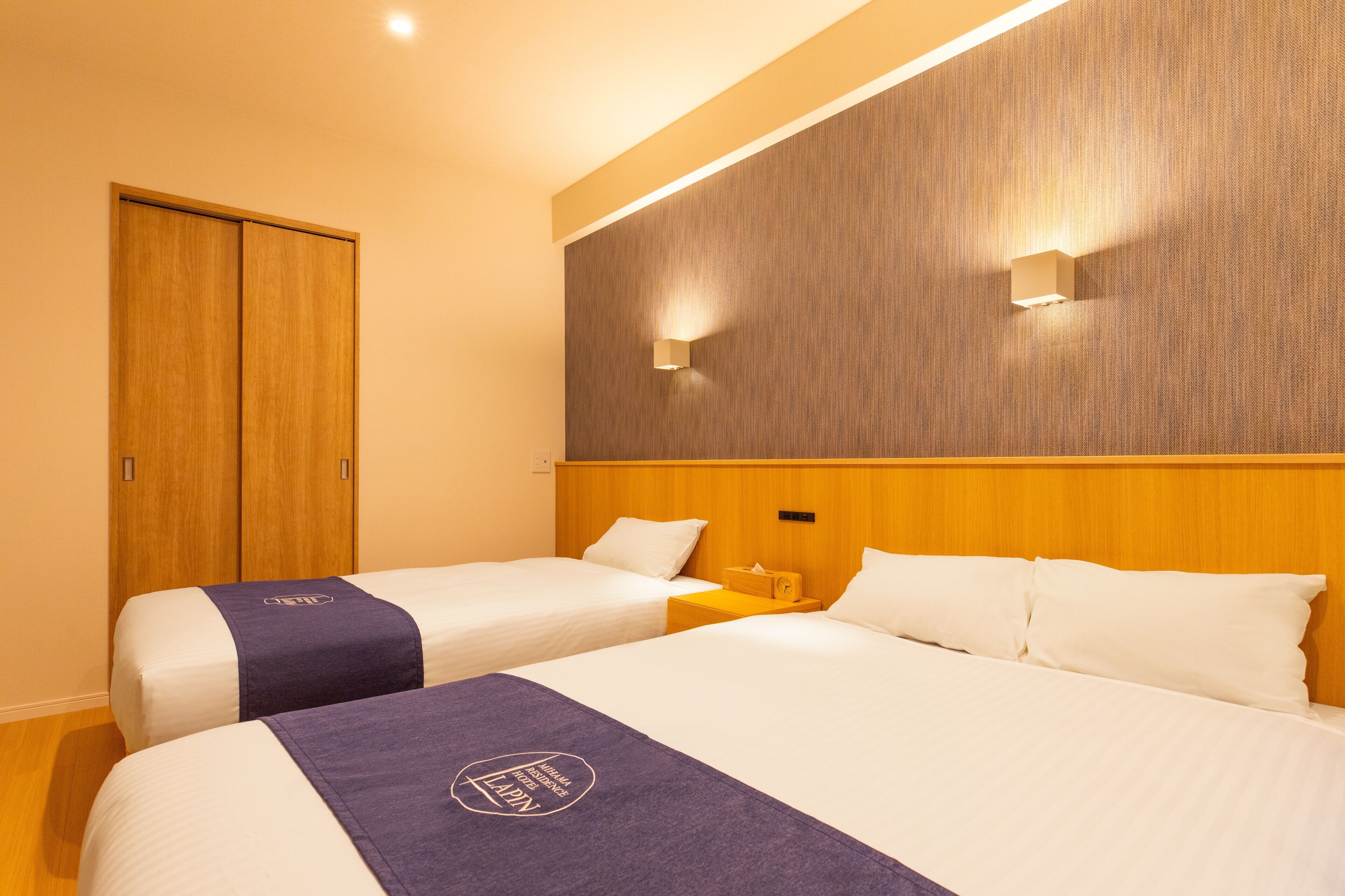 Quarto de hotel com duas camas, guarda-roupa de madeira e um painel de parede texturizado.