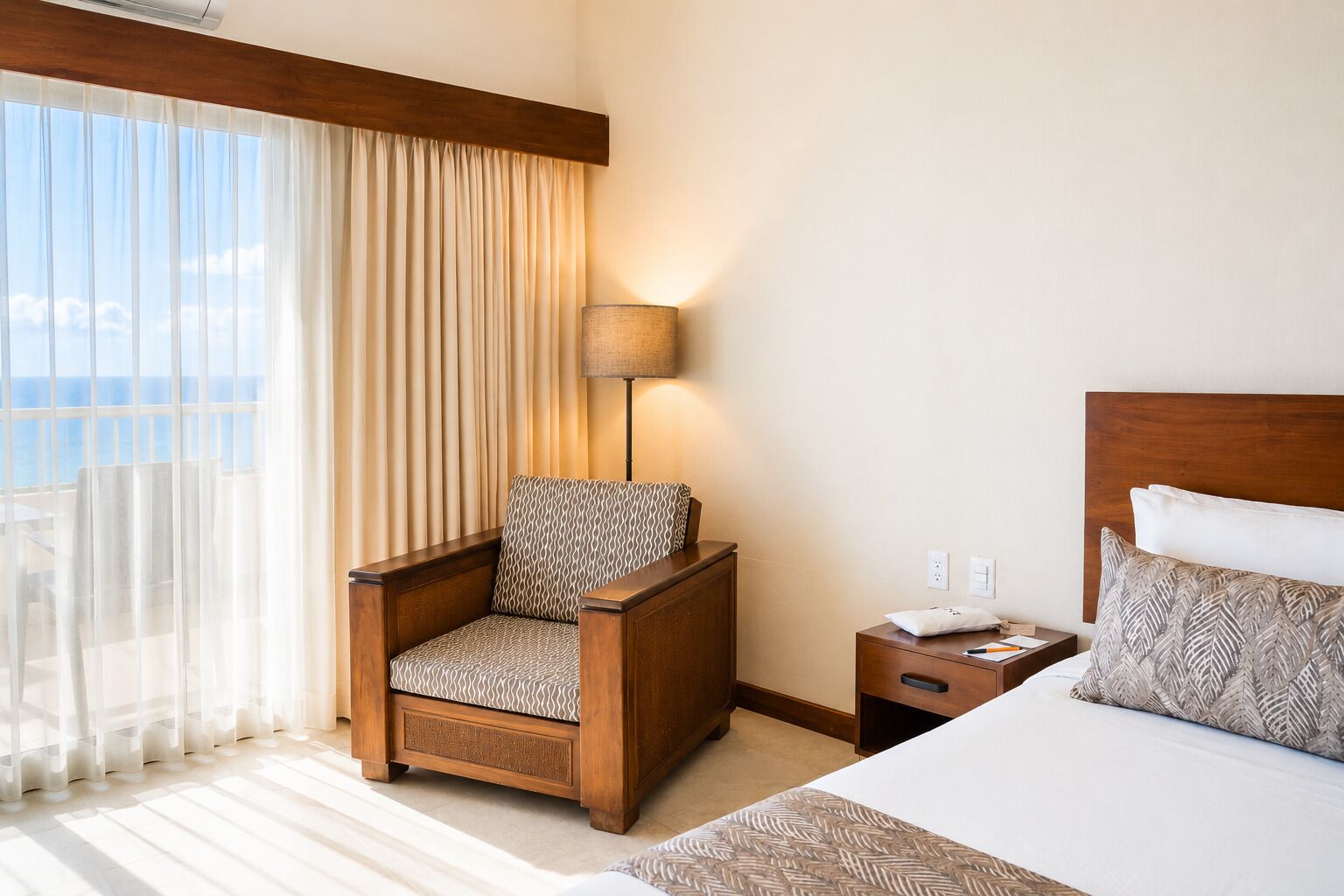 Quarto de hotel com cama, uma cadeira, mesa de cabeceira, abajur e vista para o oceano.