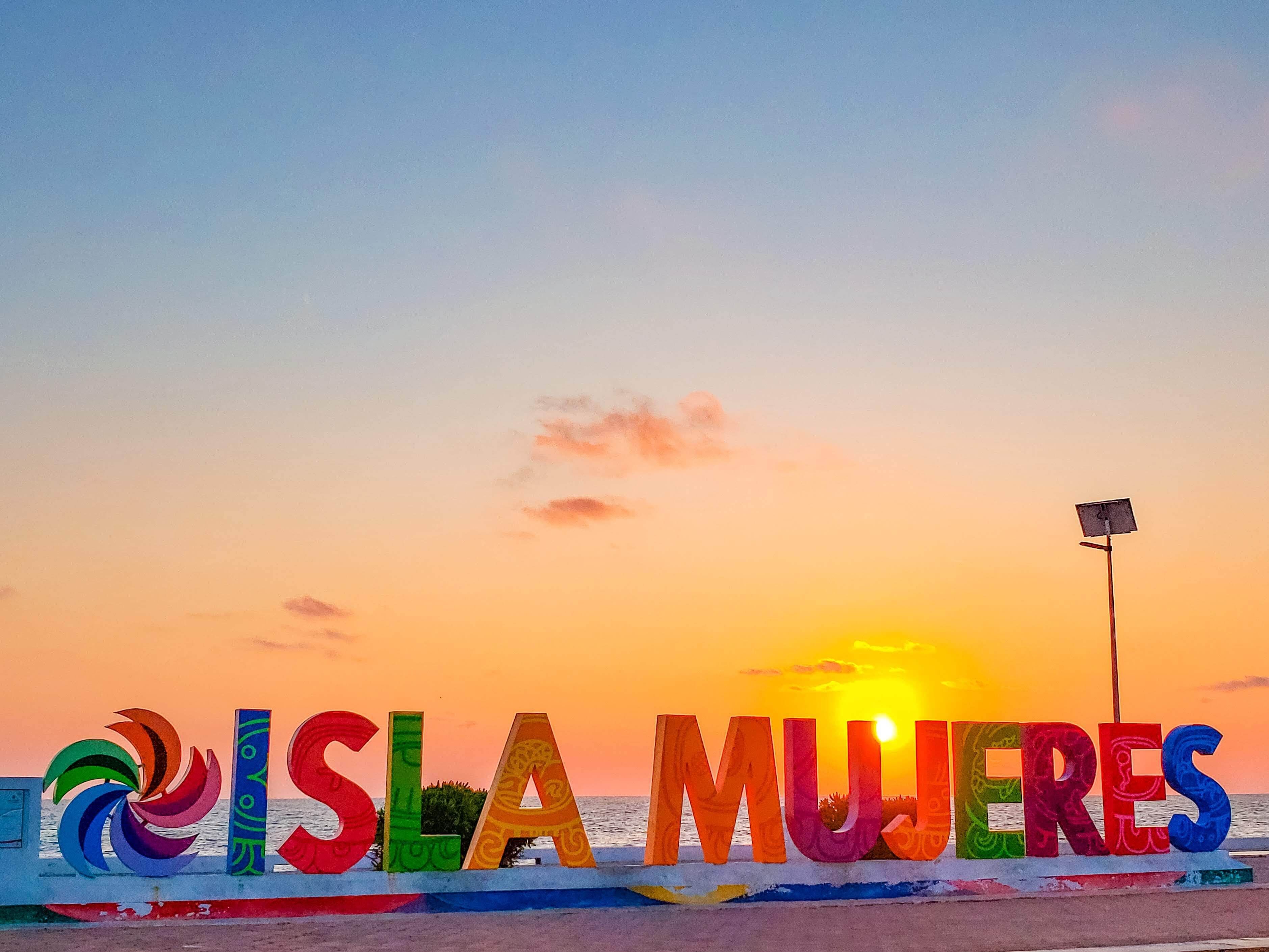 Um pôr do sol com as palavras "ISLA MUJERES" em letras coloridas sobre uma barreira.