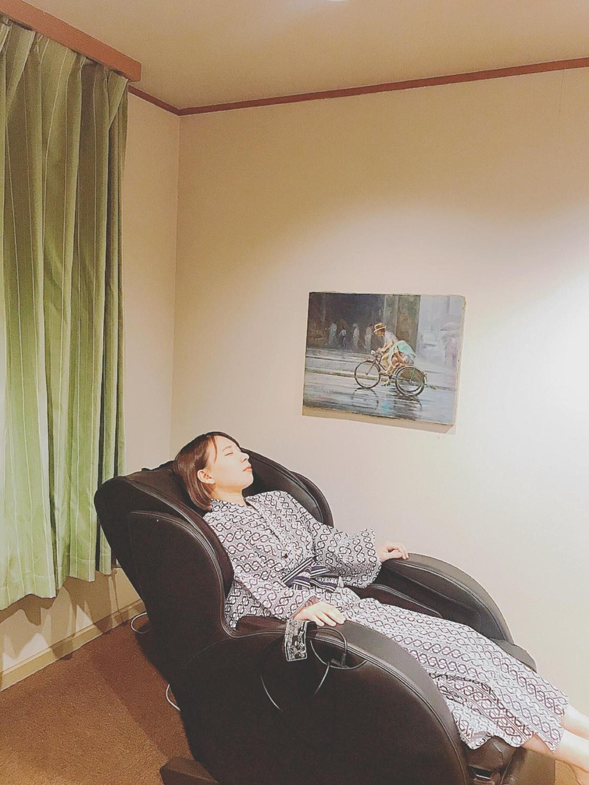 Uma pessoa relaxando em uma cadeira de massagem em um quarto com cortina verde e um quadro na parede.