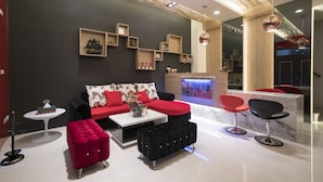 Um hall de hotel moderno com um sofá vermelho, cadeiras pretas e vermelhas, uma mesa de centro branca e um banquinho preto. Há uma mesa de recepção com balcão de mármore e uma parede decorativa com prateleiras.