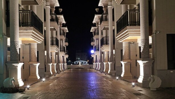 Vista noturna de um corredor de hotel com colunas e varandas, que leva a um edifício iluminado ao longe.