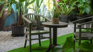 Uma área de pátio com uma mesa redonda e duas cadeiras, rodeada por diversos vasos de plantas e um pequeno tapete de grama sintética.