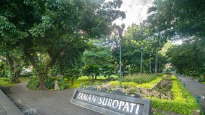 Placa com a inscrição 'TAMAN SUROPATI' em um parque com caminhos e vegetação.