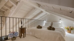 Quarto com pé direito alto e teto inclinado, cama com toalhas enroladas, mesa de cabeceira com um vaso de flores e janela com vista.