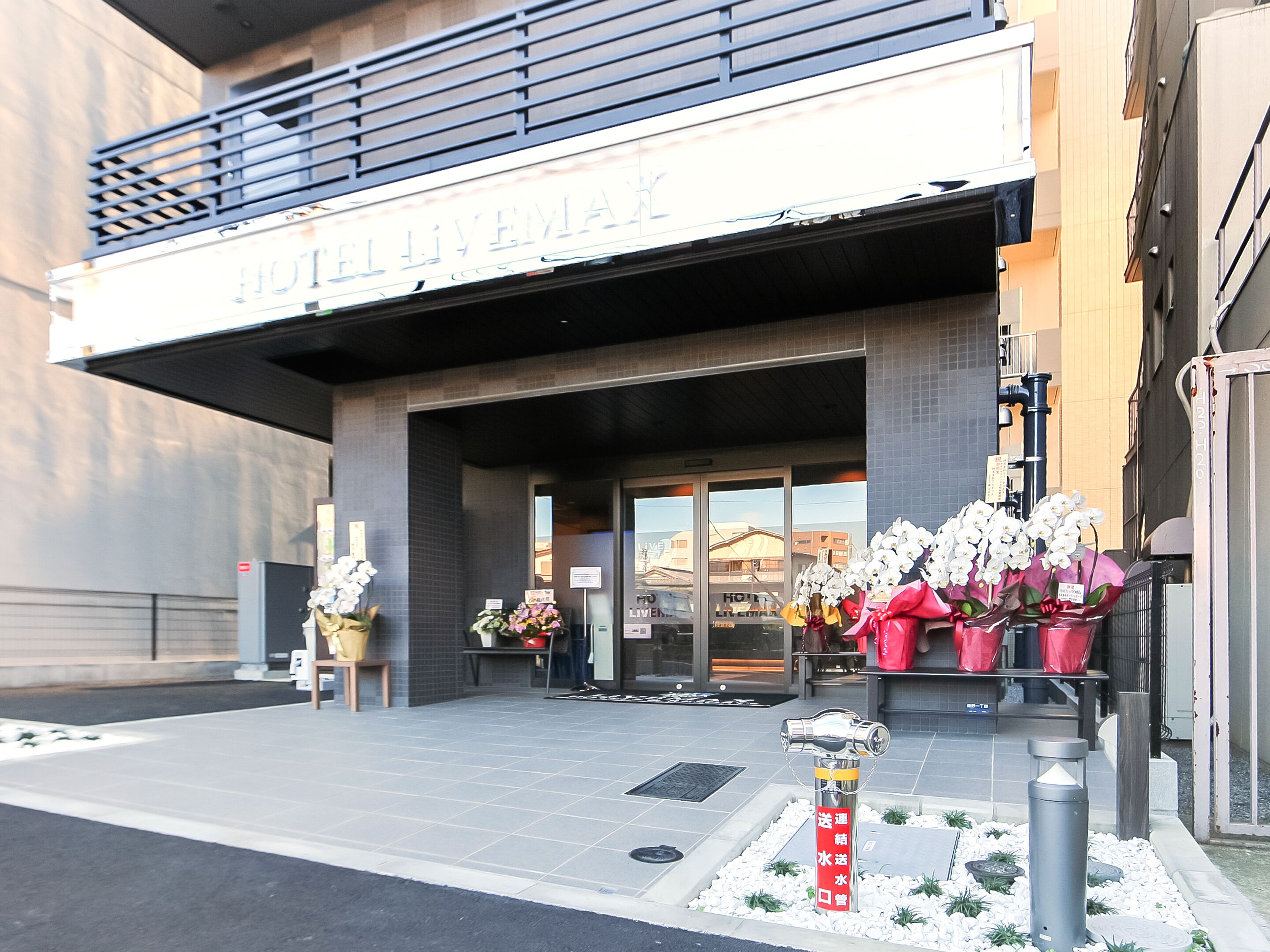 Exterior de um hotel com fachada moderna, uma placa de identificação sobre a entrada e um arranjo de flores brancas na frente.