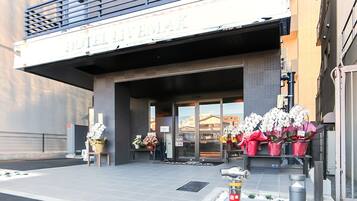 Exterior de um hotel com fachada moderna, uma placa de identificação sobre a entrada e um arranjo de flores brancas na frente.