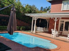 Piscina com pérgola e cadeiras, cercada por um pátio de tijolos e uma cerca branca.