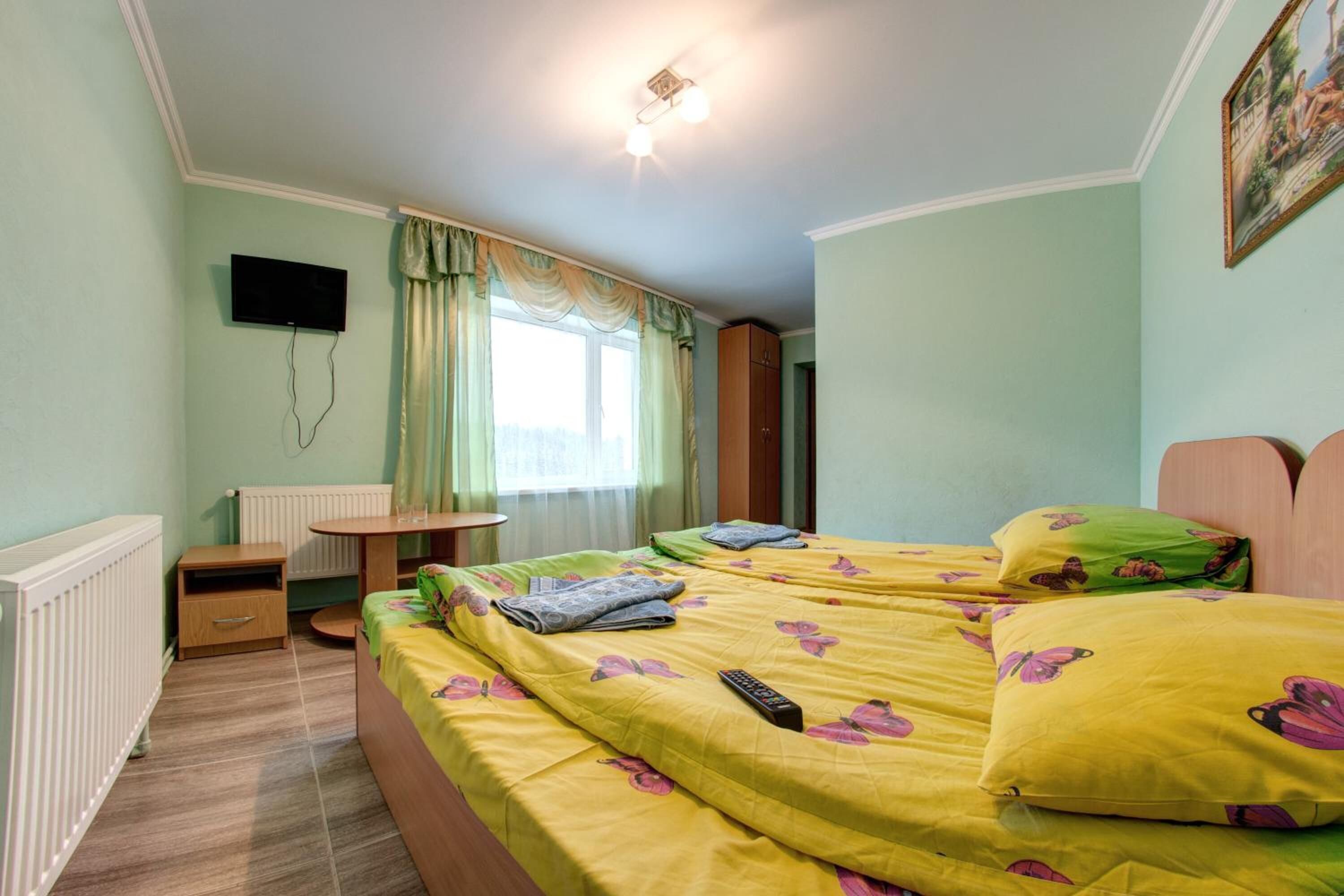 Quarto com duas camas, uma mesinha, televisão na parede e radiador.