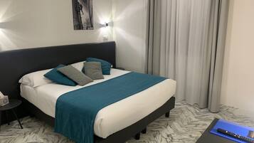 Cama bem arrumada com manta verde-água, cabeceira preta e almofadas cinzentas. Há uma pequena mesa com um controle remoto ao lado da cama.