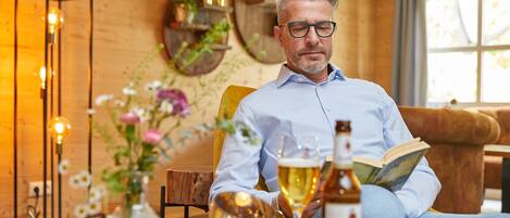 Um homem sentado num sofá lendo um livro numa sala com paredes de madeira, uma mesa com uma garrafa e copos, e um vaso com flores.