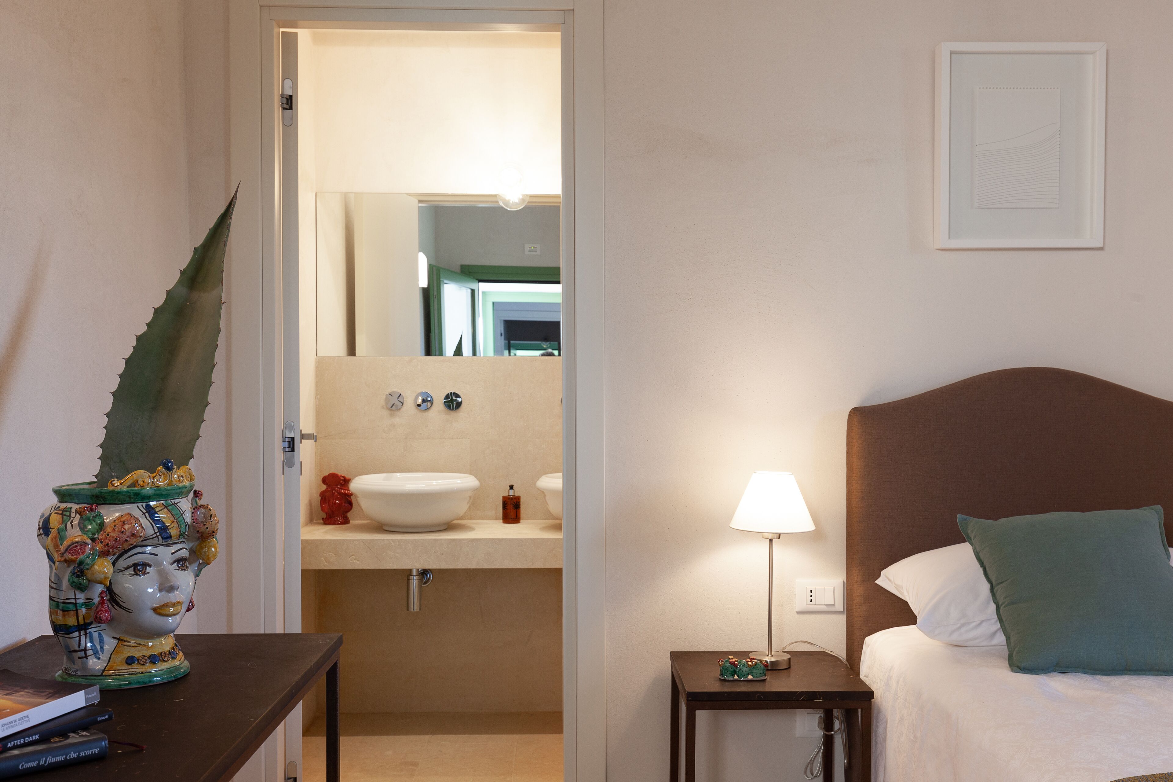 Quarto de hotel com cama, mesa de cabeceira com abajur, banheiro com pia e espelho, e um vaso decorativo.