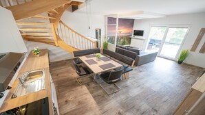 Cozinha moderna com escada de madeira, sala de jantar com mesa e cadeiras, e sala de estar com sofá e televisão.