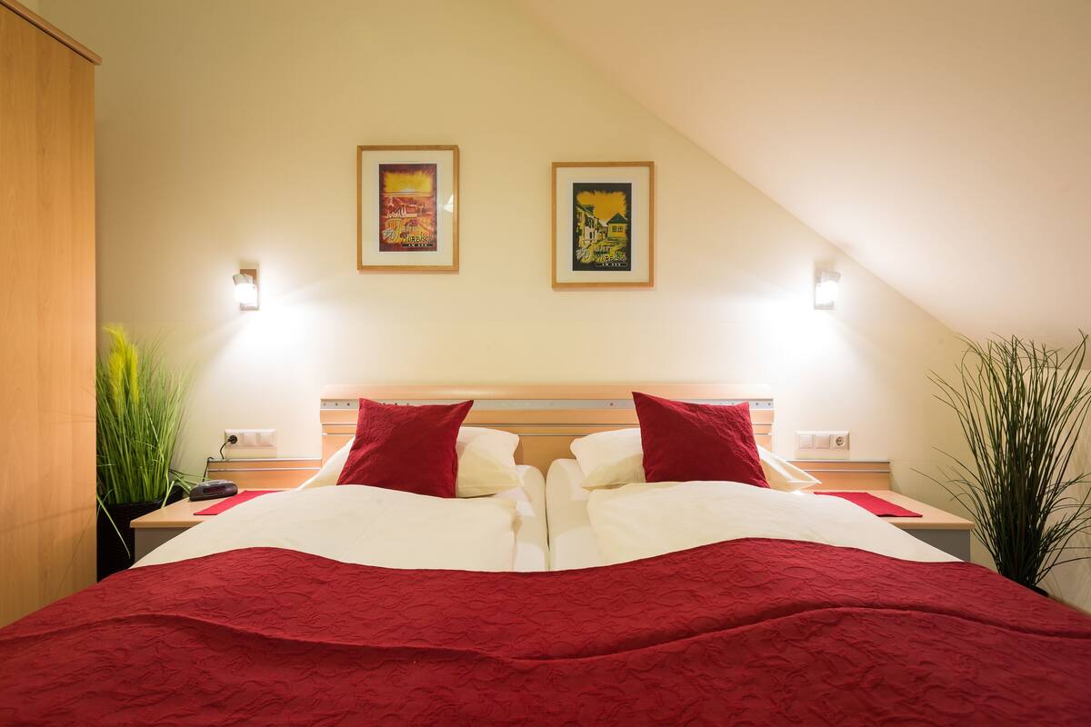 Cama bem arrumada com roupa de cama vermelha e branca, duas fotos emolduradas na parede e abajures laterais.
