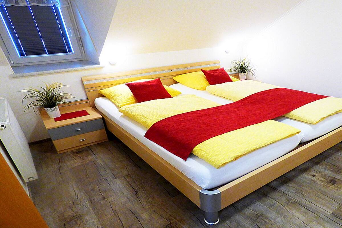 Quarto com cama de madeira, mesa de cabeceira, planta em vaso e janela com persianas.