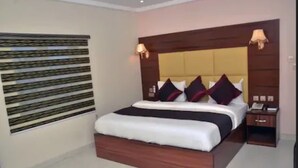 Quarto de hotel com uma cama grande, cabeceira de madeira e uma mesa de cabeceira com abajur.