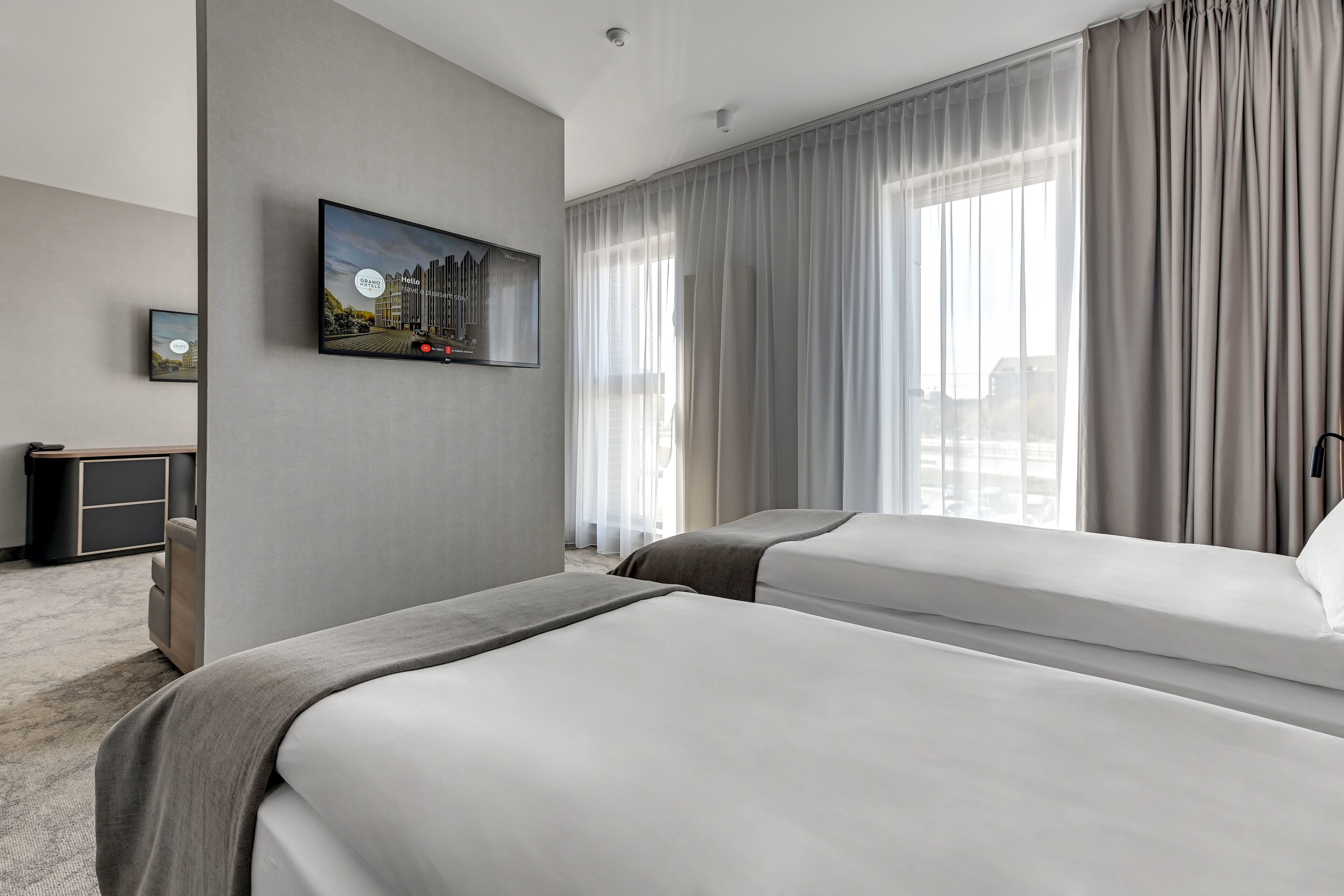 Quarto de hotel com duas camas, uma televisão de tela plana na parede e janelas amplas com cortinas.