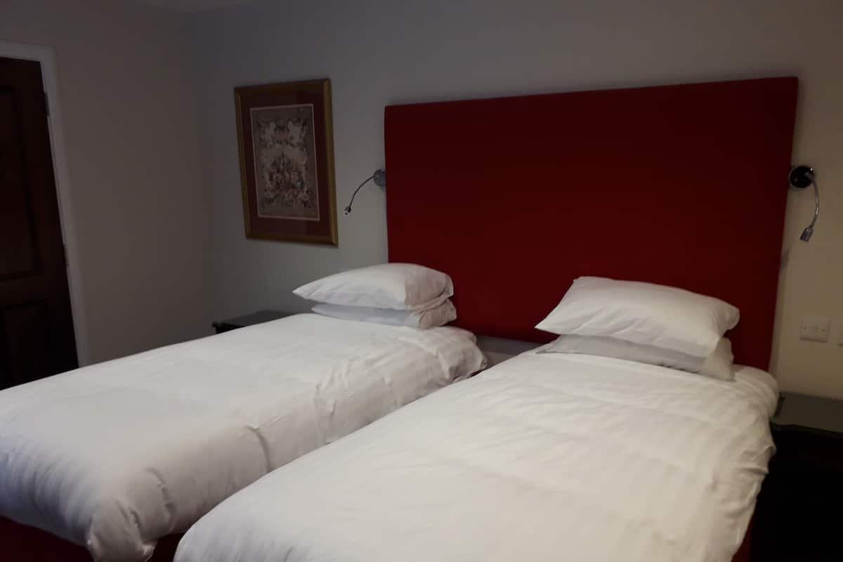 Quarto de hotel com duas camas de solteiro, cabeceira vermelha, roupa de cama branca, um quadro na parede e uma mesinha de cabeceira com abajur.
