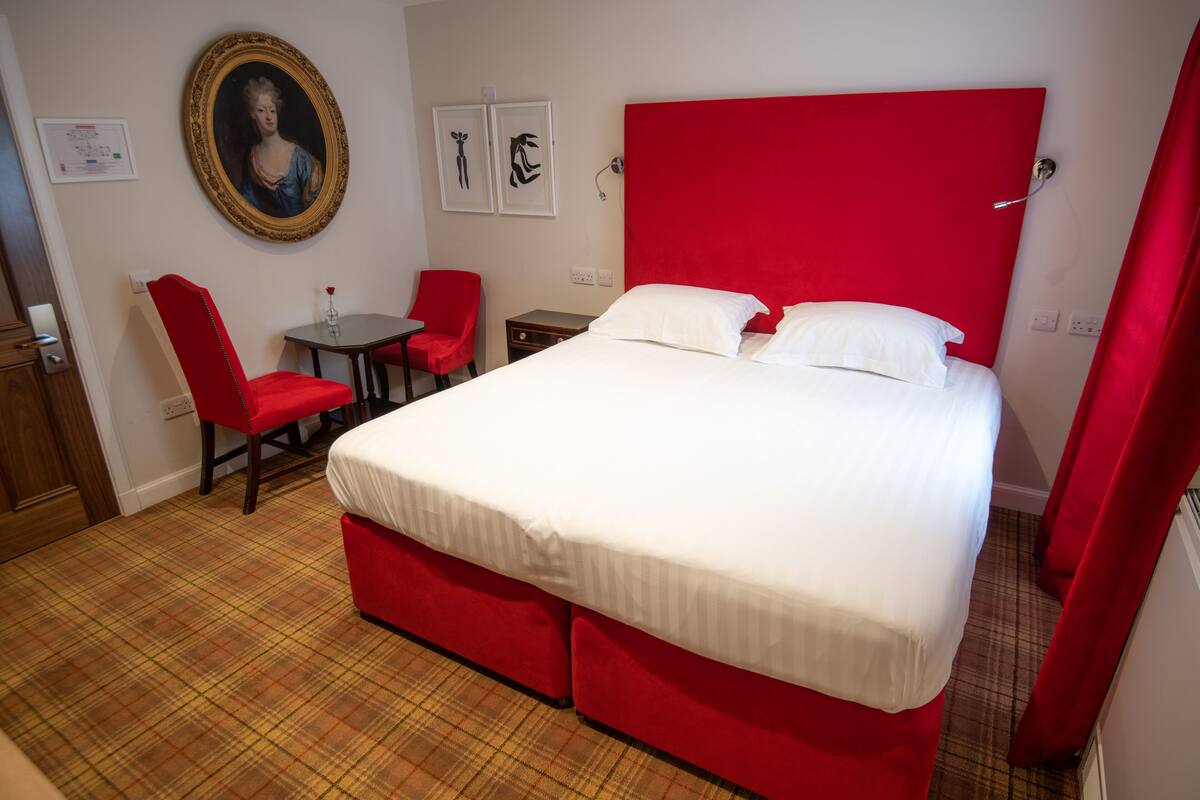 Quarto de hotel com uma cama grande, poltronas vermelhas estofadas, uma mesinha e um quadro na parede.
