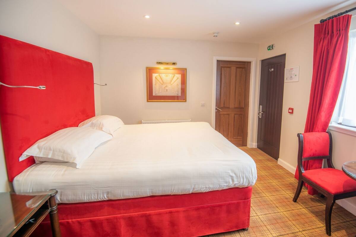 Quarto de hotel com uma cama grande, uma parede de destaque vermelha, uma porta de madeira e uma cadeira vermelha.