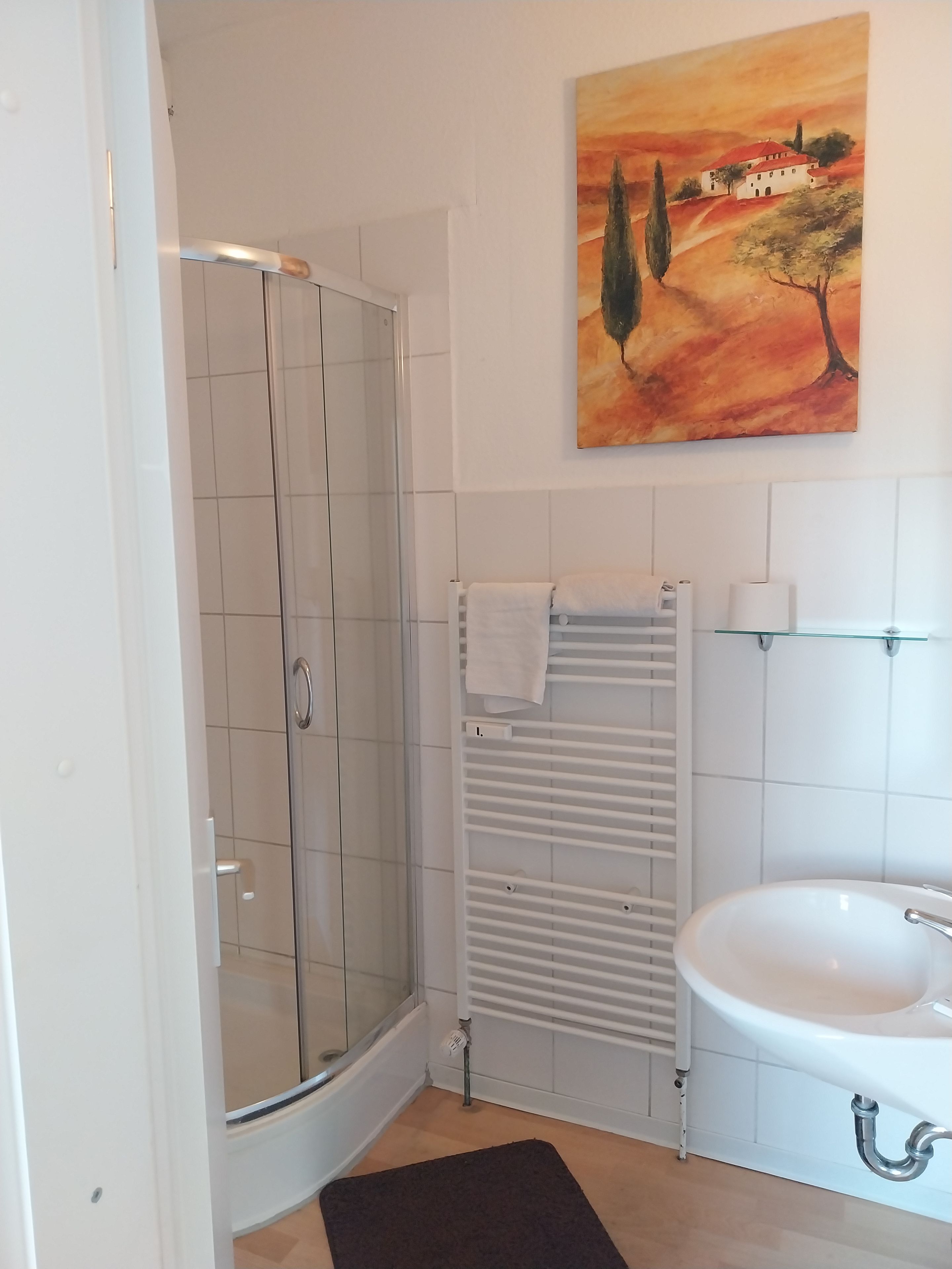 Casa de banho com chuveiro de vidro, lavatório branco e porta-toalhas.