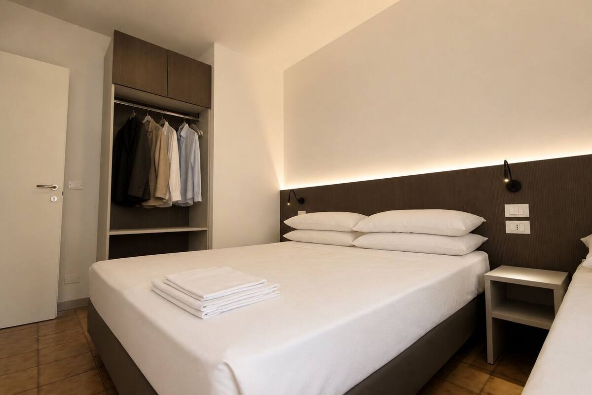 Quarto de hotel com cama, mesa de cabeceira, guarda-roupa e luminária na parede.