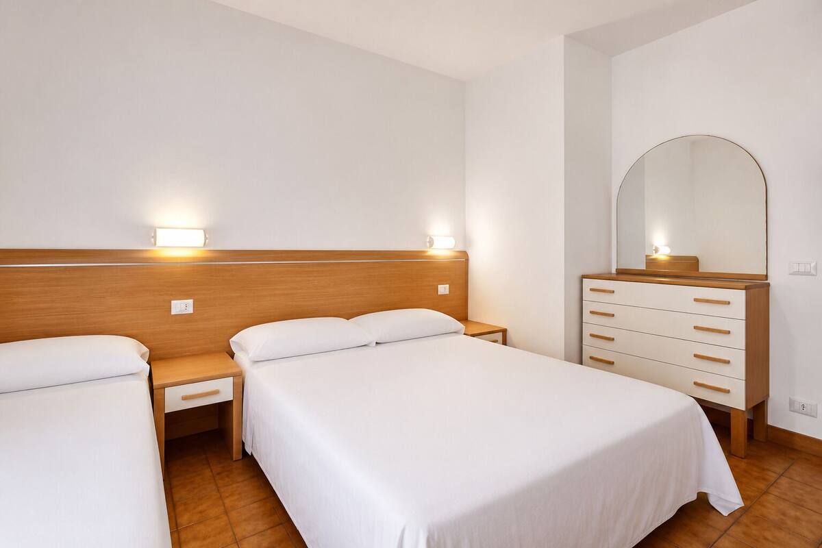 Quarto de hotel com cabeceira de madeira, duas camas de solteiro, cômoda com gavetas e um espelho.
